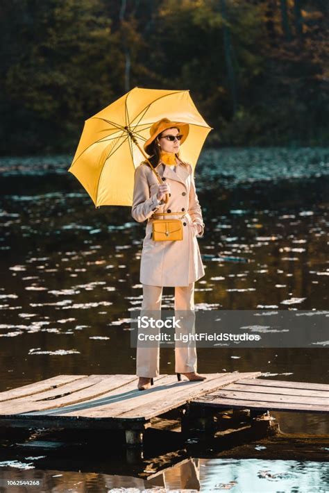 선글라스 트렌치 코트와 모자 공원에 연못 근처 노란 우산 포즈 유행 여자의 선택적 초점 가을에 대한 스톡 사진 및 기타 이미지