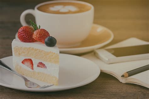 만든 맛있고 부드러운 딸기 케 잌 스마트폰와 빵집 상점 또는 카페 레스토랑에서 뜨거운 카푸치노 커피 옆에 나무 테이블에 딸기와 휘 핑된 크림 바닐라 스폰지 케이크입니다