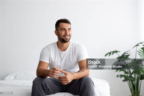 물 한 잔을 가진 미소 짓는 천년 근육질 백인 남자가 하얀 침실 내부의 빈 공간을 바라 봅니다 남자에 대한 스톡 사진 및 기타 이미지 Istock