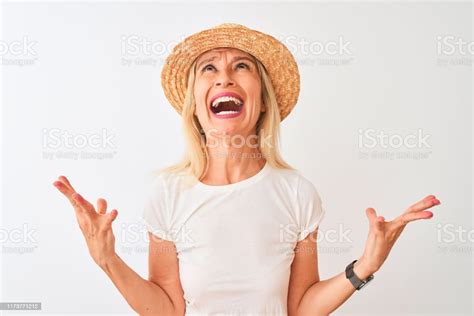 캐주얼 한 티셔츠와 모자를 입고 고립 된 흰색 배경에 서있는 중년 여성은 미친 듯이 미친 듯이 외치고 공격적인 표정과 팔을 들고