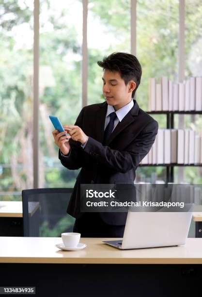휴대 전화에 타이핑 검은 양복을 입은 젊은 아시아 사업가 테이블에노트북 컴퓨터와 커피 한 잔이 있습니다 아시아에 대한 스톡 사진