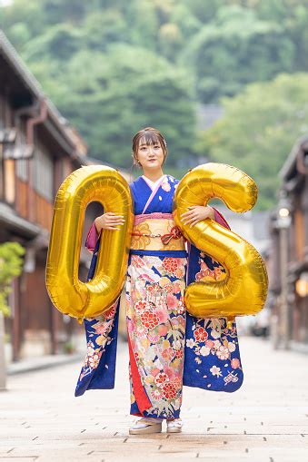 어느 여름날 파란 기모노를 입은 20세의 일본 소녀가 2와 0 모양의 황금 풍선을 들고 일본 도시의 전통적인 거리 풍경에 서 있습니다 뒤로 보기 18 19세에 대한 스톡