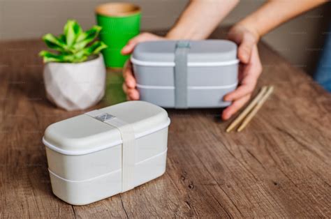 나무 테이블에 탄성 밴드가 있는 두 개의 닫힌 플라스틱 2층 도시락 배경에 도시락 하나를 들고 있는 여성 손 테이블 위의 도시락에 선택적으로 초점을 맞춥니다 사진