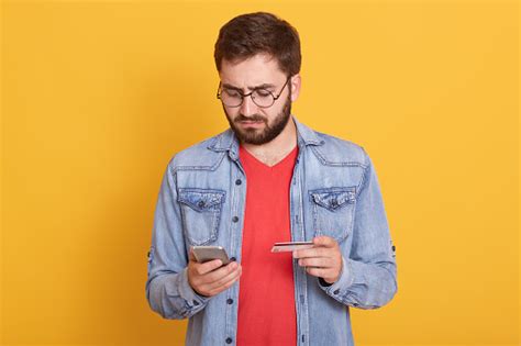 캐주얼 한 빨간 티셔츠와 데님 재킷을 입고 신용 카드와 휴대 전화를 들고 온라인 쇼핑을하고 안경을 쓰고 스튜디오에서 노란색 배경 위에 고립 된 남자의 초상화를 닫습니다 T