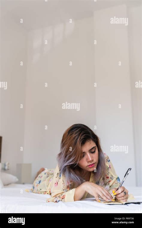 Lovely Left Handed Female Making Notes In Small Notebook While Lying On Soft Bed In Cozy Room