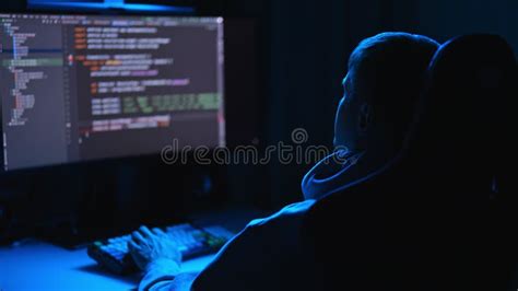 Tired Male Hacker Wearing Sweatshirt Typing On Keyboard Looking To Monitor With Text Code On