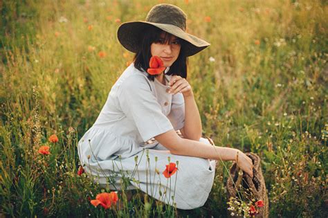 소박한 빨 대 바구니에 꽃과 일몰 빛 초원에 양 귀 비 꽃을 들고 리넨 드레스에 세련 된 소녀 여름 들판에서 편안한 모자에 보헤미안 여자 대기 모멘트 리넨에 대한 스톡 사진