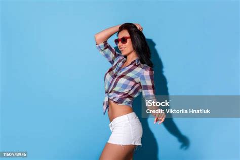 여름 시간입니다 반바지와 선글라스를 입은 여성은 파란 벽에 고립 된 맨 배를 들고 카메라를 만지는 머리를 만지는 장난스러운 측면보기로 포즈를 취합니다 스튜디오 촬영에 대한