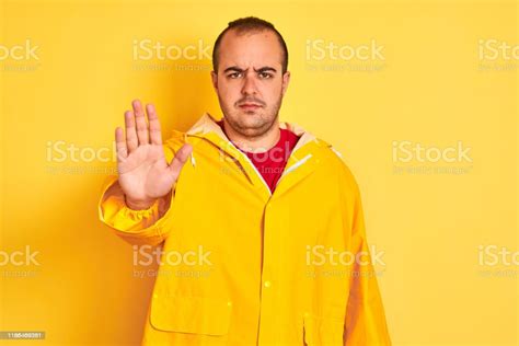 고립 된 노란색 배경 위에 서있는 비옷을 입은 청년은 손바닥으로 노래를 멈추고 있습니다 얼굴에 부정적이고 심각한 제스처로 경고 표현 가을에 대한 스톡 사진 및 기타 이미지