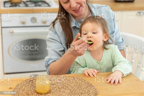 집에서 행복한 가족 부엌에 있는 숟가락으로 딸에게 먹이를 주는 어머니 지저분한 재미있는 얼굴을 가진 어린 아이는 집에서 건강에 좋은 음식을 먹는다 어린 딸에게 음식을 주는