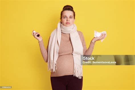 베이지 색 스웨터와 스카프를 입은 어린 아름다운 임신 한 소녀는 비강 혼잡에 대한 구제 책을 사용하며 노란 배경위에 고립 된 무기력한 표정으로 임산부를 화나게합니다 건강