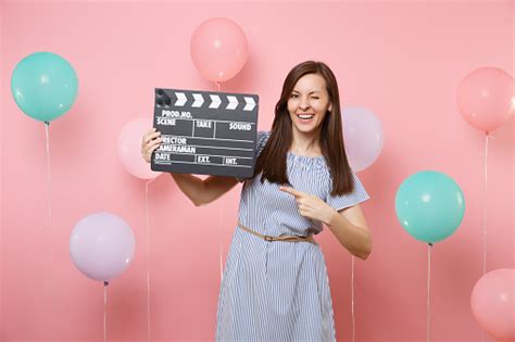 다채로운 공기 Baloons와 분홍색 배경에 Clapperboard 만드는 고전적인 흑인 영화에 지주 포인팅 검지 손가락을 점멸 하는 파란 드레스를 입고 즐거운 젊은 여자의