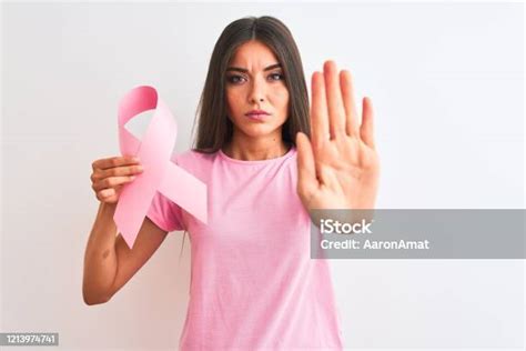 진지하고 자신감있는 표정으로 정지 표지판을하고 열린 손으로 고립 된 흰색 배경 위에 서있는 암 리본을 들고있는 젊은 아름다운 여성 방어 제스처 유방암 인식 향상 리본에 대한