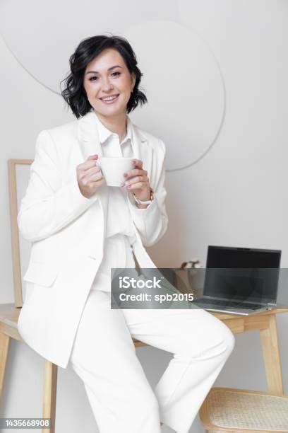 그녀의 손에 커피 머그잔을 들고 엄격한 흰색 정장을 입은 행복한 여자 미니멀리스트 인테리어의 나무 테이블에 노트북에서 작동합니다 커피 브레이크 40 44세에 대한 스톡 사진
