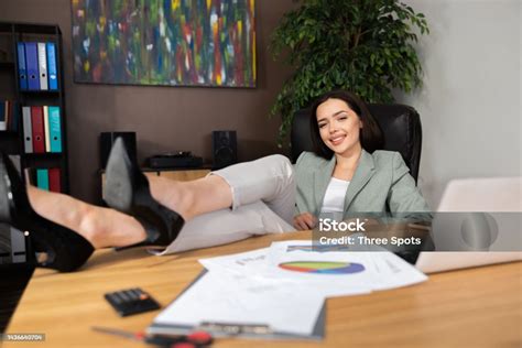 책상 위에 하이힐 신발을 들고 사무실 의자에 앉아 있는 편안한 비즈니스 여성 힘든 하루 일과를 마치고 카메라를 바라보며 웃고 있다 책상에 디지털 장치 노트북 컴퓨터 서류