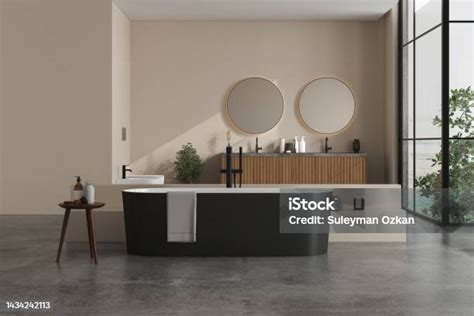 베이지 색 벽이있는 현대적인 욕실 인테리어 이중 거울이있는 대리석 분지 검은 욕조 및 회색 콘크리트 바닥 현대적인 가구가있는 미니멀리스트 욕실 나무와 가로 보기가 내려다 보