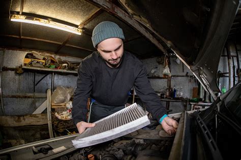 차고에서 자동차의 더러운 공기 필터를 교체하는 청년 그는 한 손에 에어 필터를 들고 다른 손은 자동차 후드에 무언가를 조이고 있습니다 측면도 와이드 샷 어안 렌즈 20 24