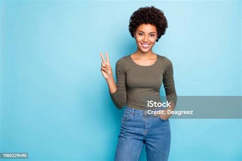 이가 귀여운 달콤한 매력적인 아프리카 여자 친구의 사진은 파란색 선명한 색상 배경 위에 자신있게 고립 된 주머니에 손을 들고 청바지 데님을 입고 V사인을 보여주는 갈색에