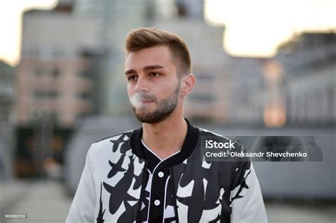 흰색과 검은 색 양복을 입은 잘 생긴 남자가 담배의 연기를 내뿜습니다 세련된 양복을 입고 담배를 피우는 그루터기와 모델 헤어 스타일을 가진 남자 25 29세에 대한 스톡