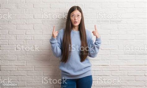 흰색 벽돌 벽을 통해 젊은 Chinise 여자 놀라게 했 고 올려 손가락으로 가리키는 그리고 팔 놀 랐 어 요 가리키기에 대한 스톡 사진 및 기타 이미지 Istock