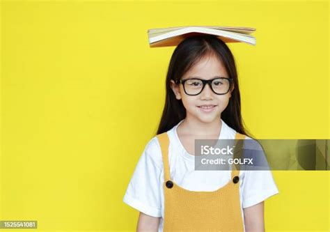 웃고 있는 아시아의 어린 소녀는 머리에 책이 있는 안경을 쓰고 복사 공간이 있는 노란색 배경에 고립된 카메라를 보고 있다 학생과 숙제 개념 4 5세에 대한 스톡 사진 및