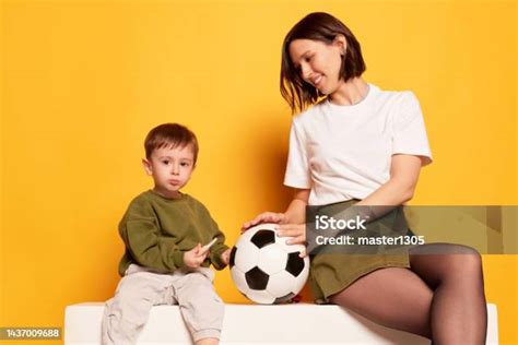 밝은 노란색 배경에 고립된 축구 공을 들고 있는 귀여운 어린 아들과 함께 웃고 있는 젊은 어머니 가족 사랑 모성 및 어머니의 날