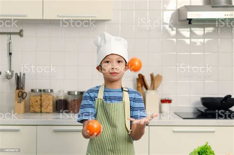 앞치마와 요리사 모자를 쓴 행복한 웃는 아시아 소년의 초상화 오렌지 과일을 들고 부엌에 서서 공중에 던지는 귀여운 어린이 요리사는 혼자 음식을 요리하는 것을 즐기고 아이는