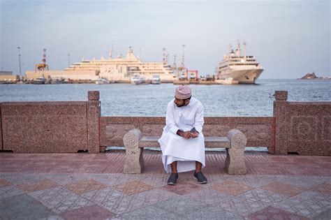 무스카트의 코니쉬 벤치에 앉아 바다물과 유람선을 배경으로 앉아 전통 무슬림 의상을 입은 젊은 무슬림 오만 남자 가정 생활에 대한