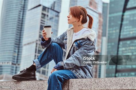 그녀의 얼굴에 문신을 가진 세련 된 빨간 머리 소식통 소녀 그녀의 얼굴이 흐린 아침에 모스크바의 고층 빌딩 앞에 앉아 테이크아웃 커피를 들고 데님 코트를 입고 가을에 대한