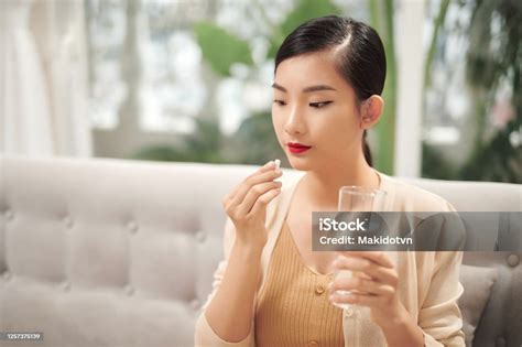 집에서 옷을 입은 아픈 아름다운 소녀가 물 한 잔으로 창문 근처에서 약을 복용합니다 건강관리와 의술에 대한 스톡 사진 및 기타 이미지 건강관리와 의술 건강한 생활방식