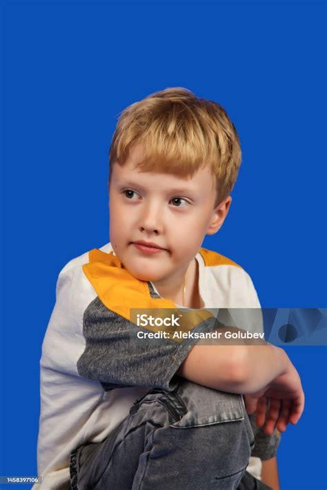 흰색 티셔츠를 입은 웃는 어린 소년이 파란색 고립된 배경에 앉아 시선을 돌린다 스튜디오에서 포즈를 취하는 캐주얼한 옷을 입은