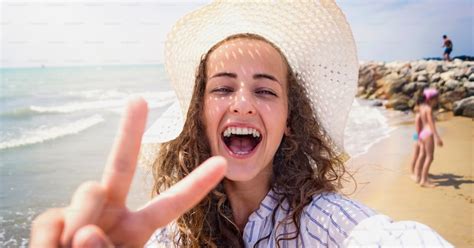 줄무늬 파란색과 흰색 셔츠와 모자를 쓰고 해변에서 아름다운 젊은 여성이 웃고 셀카를 찍고 평화의 표시를 보여줍니다 해변에서 즐거운 시간을 보내십시오 사진