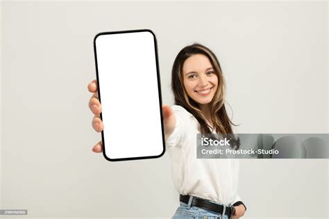 흰 블라우스를 입은 웃는 젊은 갈색 머리 여성이 빈 화면으로 스마트 폰을 제시합니다 보여주기에 대한 스톡 사진 및 기타 이미지 보여주기 여자 전화 Istock