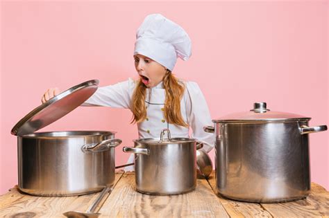 분홍색 스튜디오 배경에 고립 된 큰 프라이팬이있는 어린이 부엌에서 흰색 요리 유니폼과 거대한 요리사의 모자를 입은 어린 소녀 6 7 살에 대한 스톡 사진 및 기타 이미지