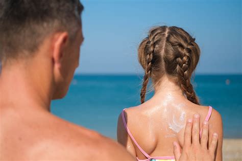 아버지는 해변에서 딸의 등에 보호 크림을 바르고 있습니다 한 남자가 아이의 몸에 자외선 차단제 로션을 들고 있다 바다에 의해 자외선 차단제를 가진 귀여운 어린 소녀 공간을