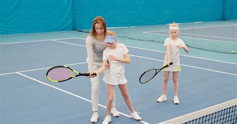 운동복을 입은 젊은 테니스 트레이너가 경기장에서 개별 훈련 중에 그녀와 상담하는 동안 두 명의 귀여운 소녀 중 한 명이 들고 있는 라켓을 들고 있습니다 사진