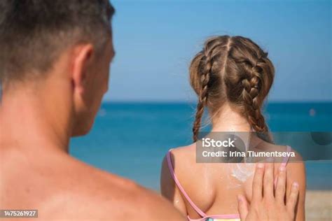 아버지는 해변에서 딸의 등에 보호 크림을 바르고 있습니다 한 남자가 아이의 몸에 자외선 차단제 로션을 들고 있다 바다에 의해