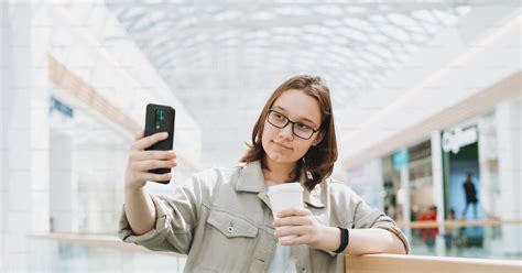 공공 장소 쇼핑몰에서 종이 커피 컵으로 셀카를 찍는 휴대 전화를 사용하는 안경을 쓴 젊은 갈색 머리 십대 소녀 사진 Unsplash의 셀카 이미지