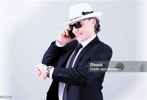 성공으로 가는 길에 비즈니스 라이프 양복을 입고 흰 모자를 쓰고 선글라스를 착용하고 스마트폰으로 대화를 나누며 미소를 지으며 하얀 배경을 배경으로 시계를 바라보고 있다