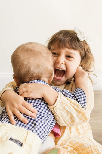 3 살짜리 유아 소녀가 9 개월 된 팬데믹 아기 형제를 즐겁게 들고 Suspensioners 및 밝은 노란색 바지가 달린 Navy White 격자 무늬 버튼 다운 셔츠를