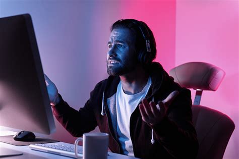 컴퓨터 모니터를보고 다채로운 배경에 앉아있는 동안 몸짓을하는 좌절 한 청년 게이머에 대한 스톡 사진 및 기타 이미지 게이머