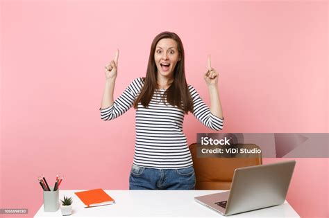 젊은 여 자가 일을 집게 손가락을 가리키는 파스텔 핑크 배경에 고립 된 Pc 노트북과 흰색 책상 근처에 서 흥분 성과 비즈니스 경력 개념입니다 광고에 대 한 공간을 복사