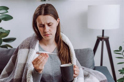 열이 나는 건강하지 못한 화가 난 여자는 높은 온도의 체온계를 보고 있다 아픈 소녀는 독감이나 감기에 걸리고 코로나19 증상이 있으며 아픈 여성은 집 소파에 담요를 덮고