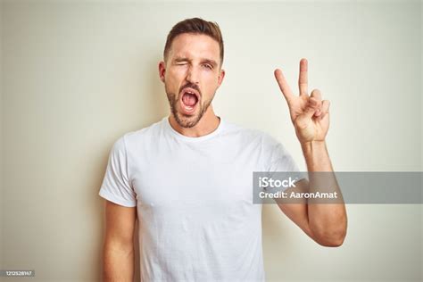 고립 된 배경 위에 캐주얼 한 흰색 티셔츠를 입은 젊은 잘생긴 남자는 승리의 표지판을하고 있는 카메라에서 행복한 얼굴 윙크와