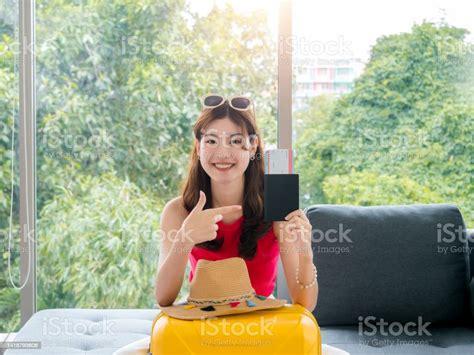 선글라스를 착용 한 젊은 행복한 아시아 여성은 여권과 항공권을 웃으며 보여 주며 해변 모자 노란색 여행 가방 및 유리 창 근처의 소파에 앉아 여름 휴가 컨셉으로 여행 할