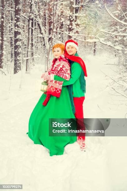 젊은 부부 남자와 여자 남편과 아내는 산타의 도우미 가슴 가득 선물 및 거 대 한 사탕으로 눈 아래 겨울 숲에서의 엘 프의 전형적인 꽃의 의상에서 걷고 있다 2명에 대한
