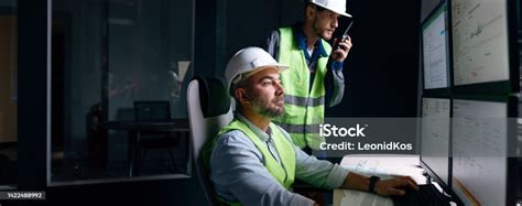 공장에서 안전 조끼와 하드 모자를 착용 한 주요 엔지니어 및 근로자 운영자는 Scada 시스템 및 Industry 40을 사용합니다 두 명의 연산자가 Ui가있는 화면을