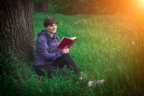 Маленькая девочка читая книгу сидя на траве около дуба на заходе солнца ...