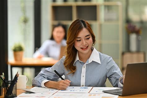 한 젊은 전문직 종사자가 노트북과 펜을 사용하여 책상에서 차트와 그래프를 검토하면서 분석 및 멀티태스킹 기술을 보여주며 미소를 짓고 있습니다 20 24세에 대한 스톡 사진