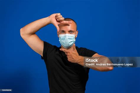 파란 눈을 가진 젊은 백인 남자는 손으로 손가락 프레임을 만드는 고립 된 파란색 배경 위에 서 의료 마스크를 착용 창의력과 사진 개념 건강한 생활방식에 대한 스톡 사진 및